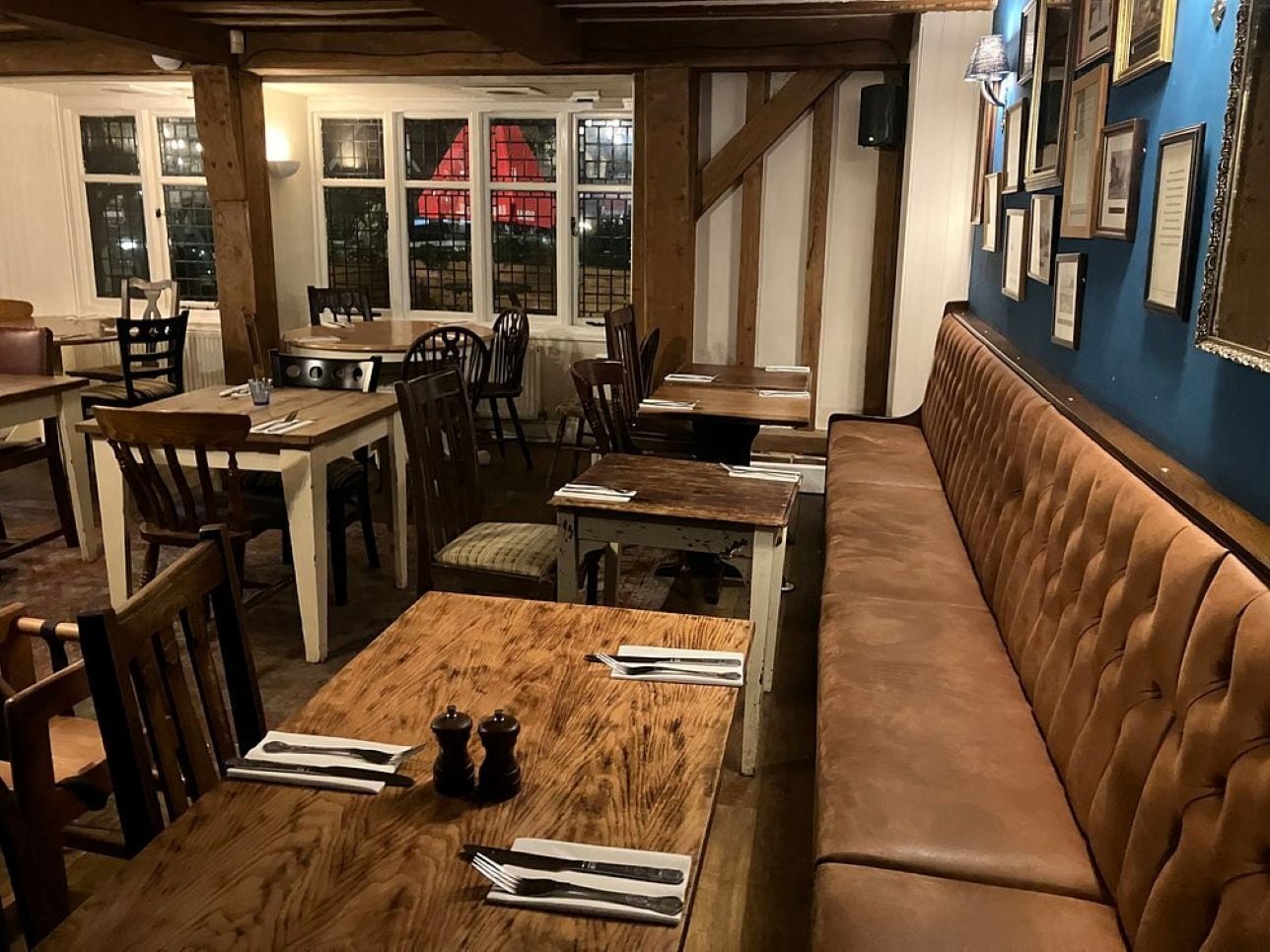 Um restaurante acolhedor com mesas de madeira, um sofá de couro, quadros nas paredes e janelas amplas com vidro avermelhado.
