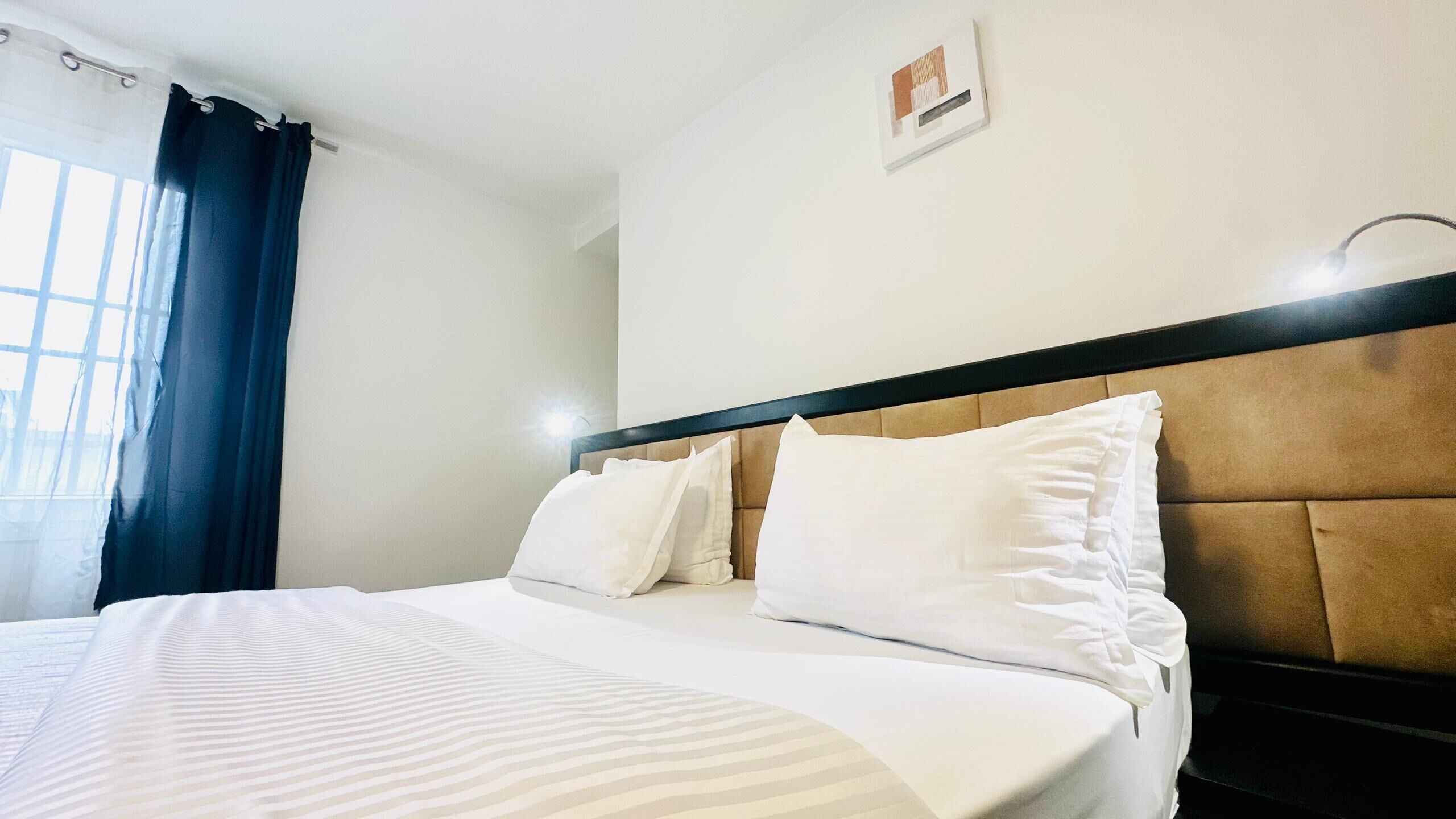 Cama bem arrumada com lençóis brancos e manta listrada, uma cabeceira escura e uma janela com cortinas.