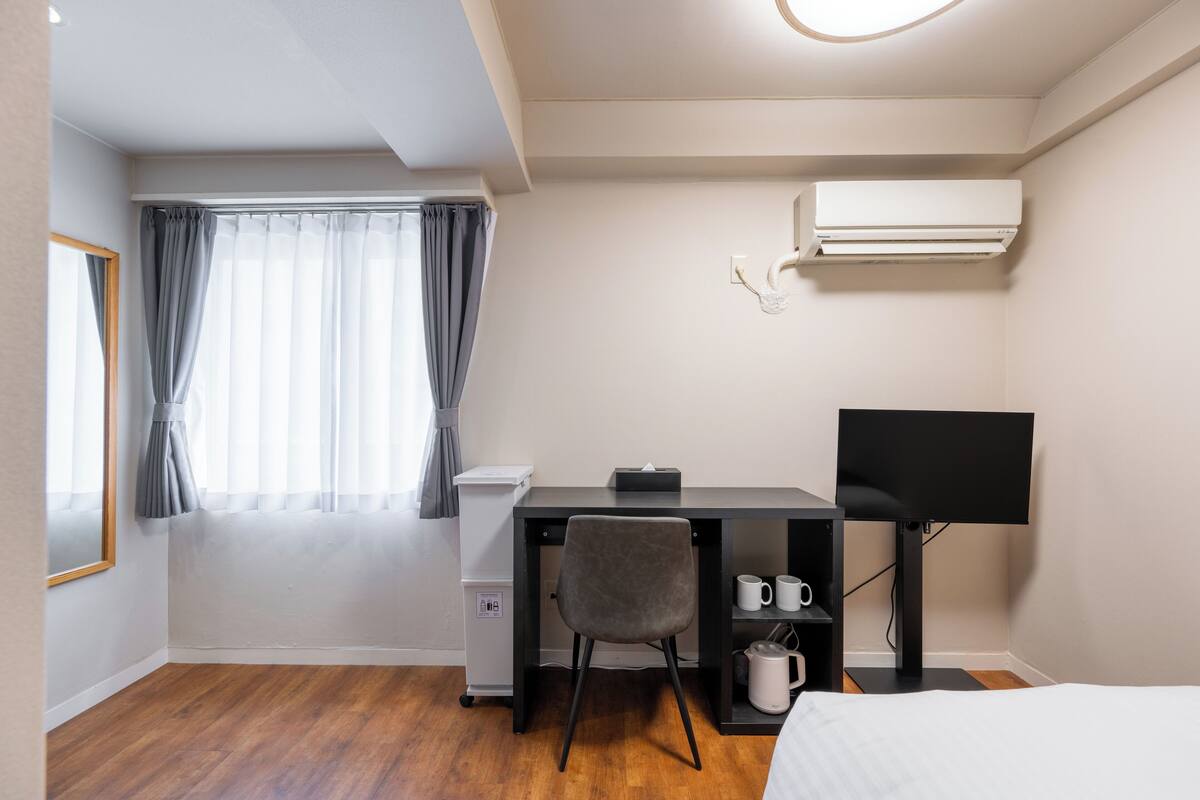 Quarto de hotel com piso de madeira, uma escrivaninha com cadeira, televisão, cafeteira e ar-condicionado.