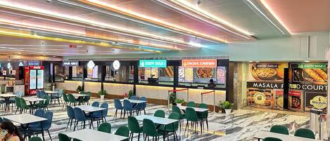 Um restaurante com cadeiras e mesas verdes, uma geladeira Coca-Cola e balcões de comida com placas indicando 'UDUPI DOSA' e 'CHINA CROWN'.
