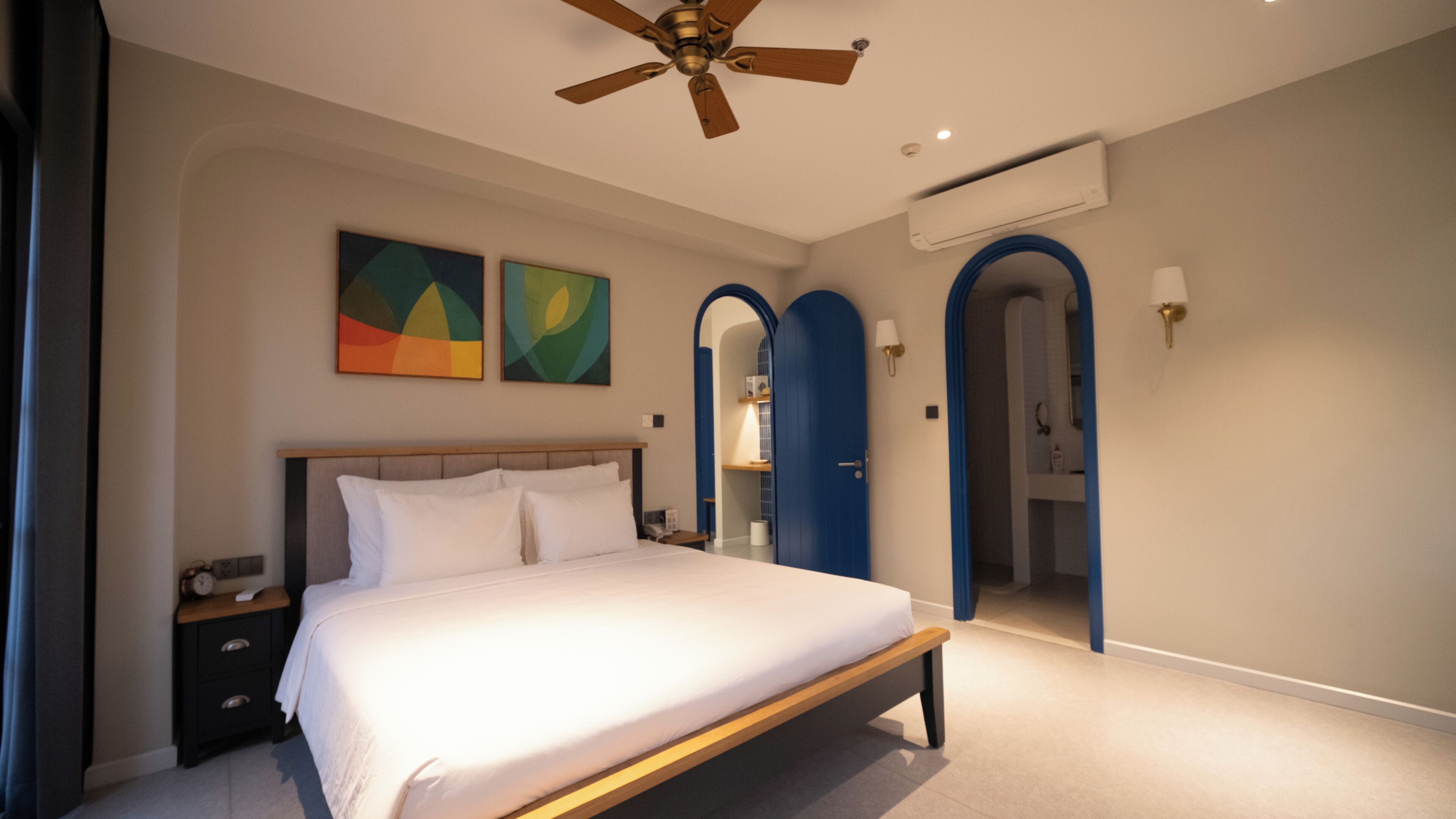 Quarto com uma cama, ventoinha no teto, duas pinturas, mesa de cabeceira, porta azul e ar condicionado.