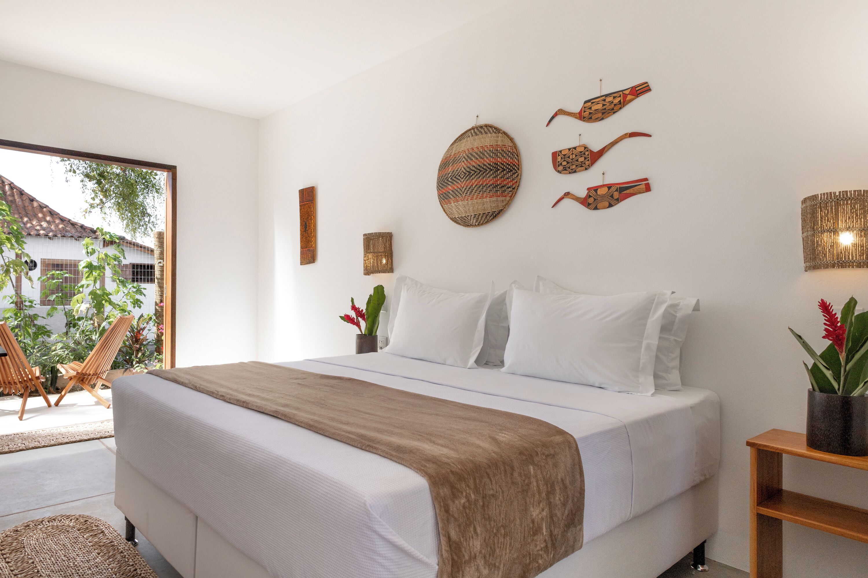 Quarto com cama grande, roupa de cama branca, manta marrom e vista para área externa com cadeiras e plantas.