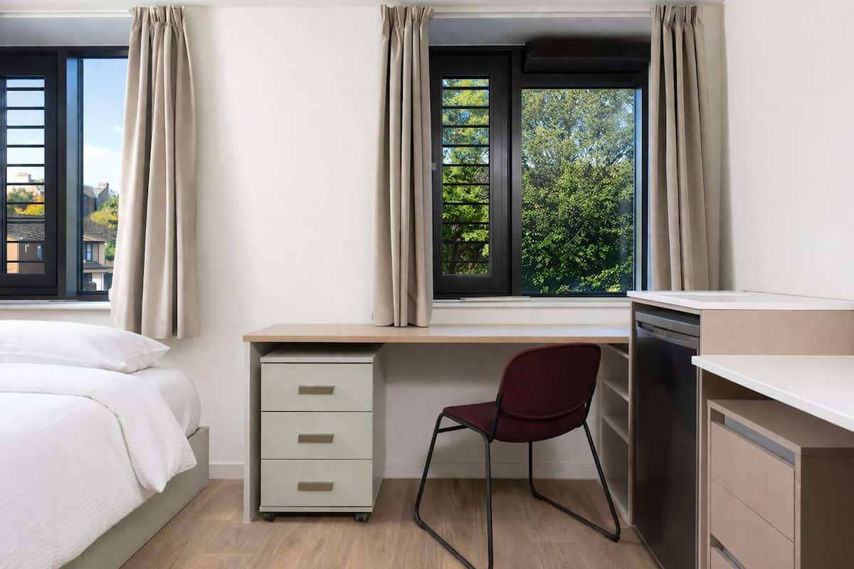Um quarto com cama, escrivaninha, cadeira e janela com cortinas.
