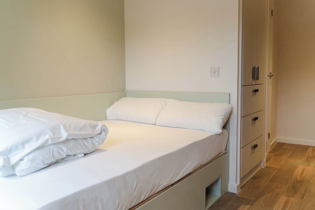 Cama bem arrumada com lençóis brancos em quarto com piso de madeira e parede branca.