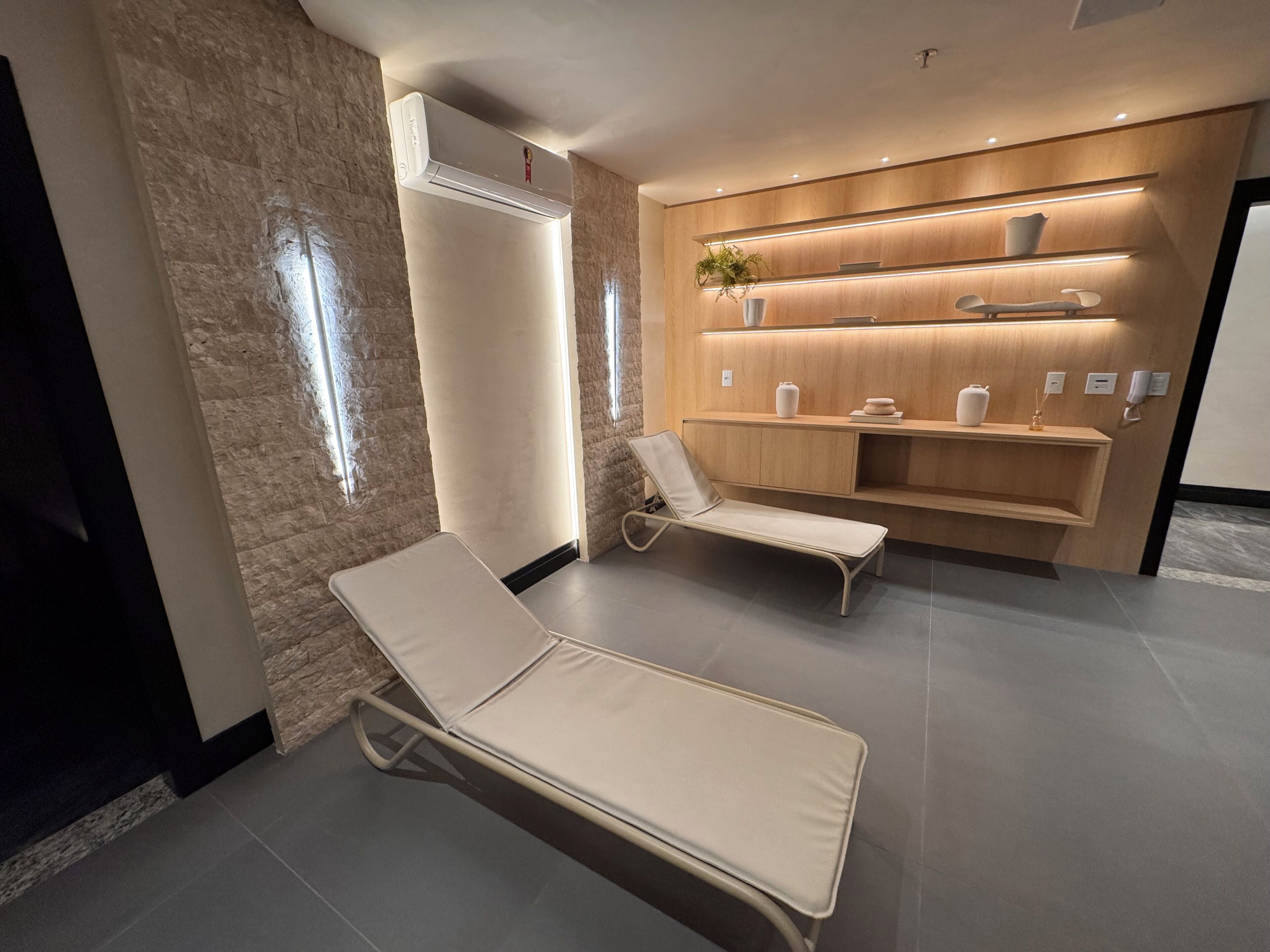Um quarto de spa moderno com uma poltrona de descanso, um ar condicionado fixado na parede e uma estante com objetos decorativos.