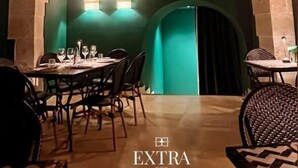 Um espaço de refeições em ambiente interno com um arco de pedra, mesas postas com copos e talheres, e uma parede verde ao fundo.