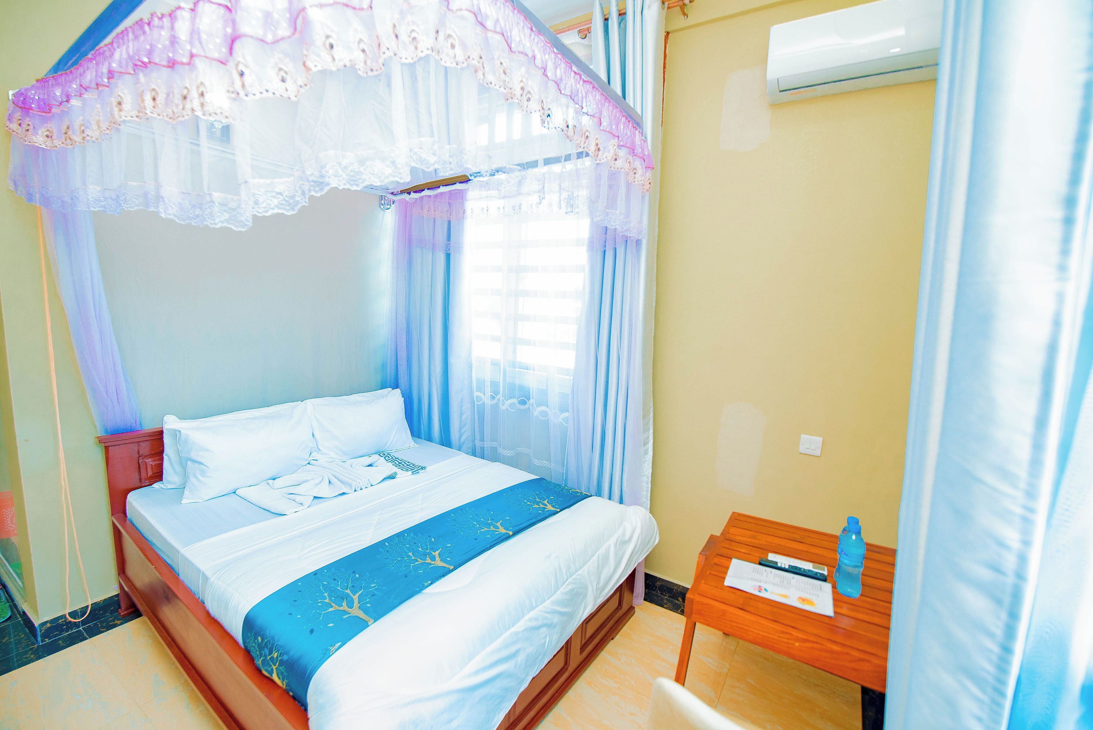 Uma cama bem arrumada com colcha azul e branca, uma mesa de cabeceira de madeira com uma garrafa de água e uma janela com cortinas leves.