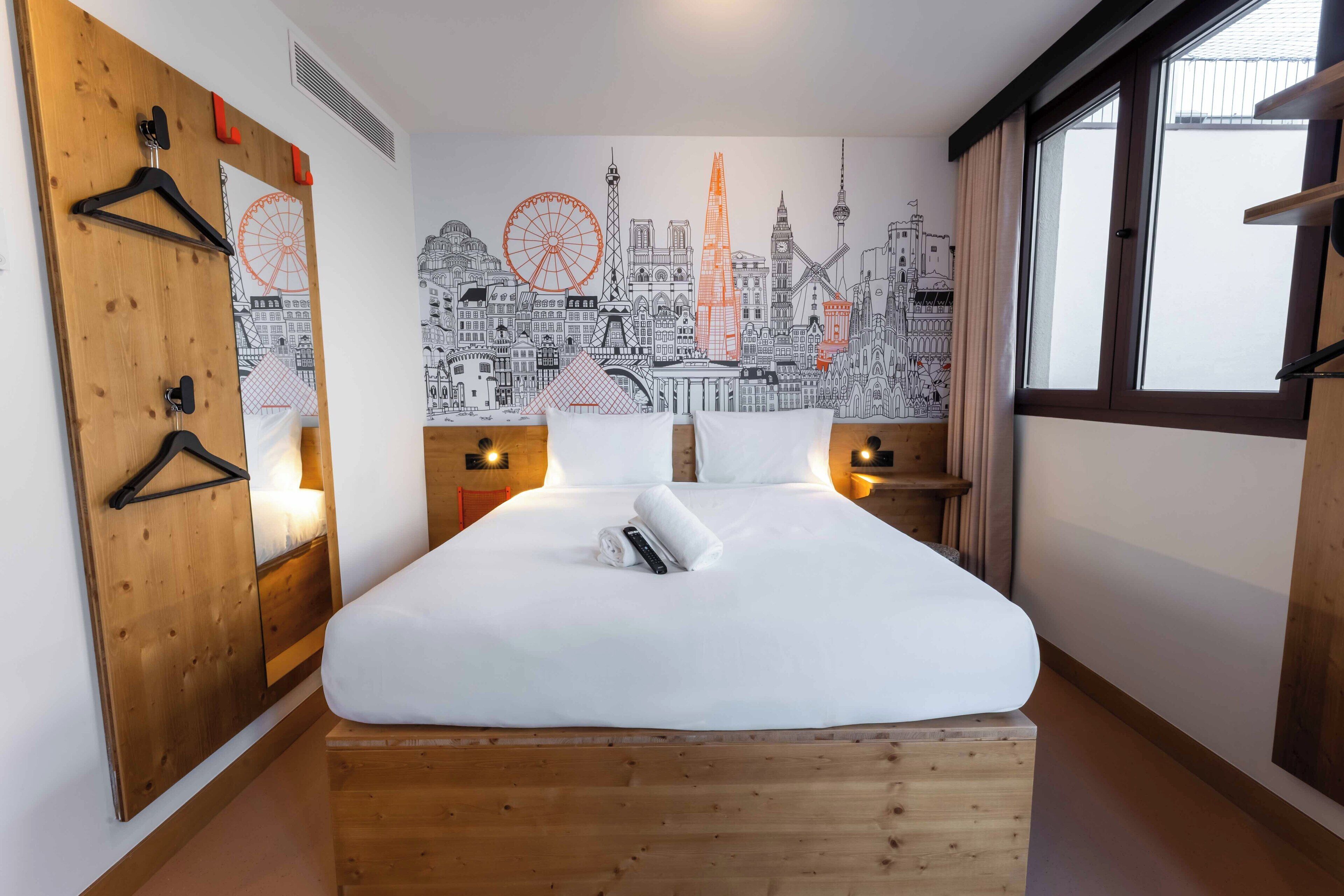 Quarto de hotel com cama grande, cabeceira de madeira, um mural com vista urbana, guarda-roupa de madeira e janela com cortinas.