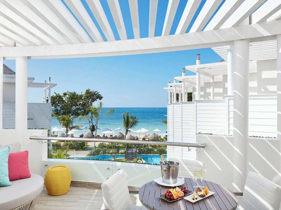Uma varanda com vista para o oceano, piscina e uma mesa com bebida e frutas.