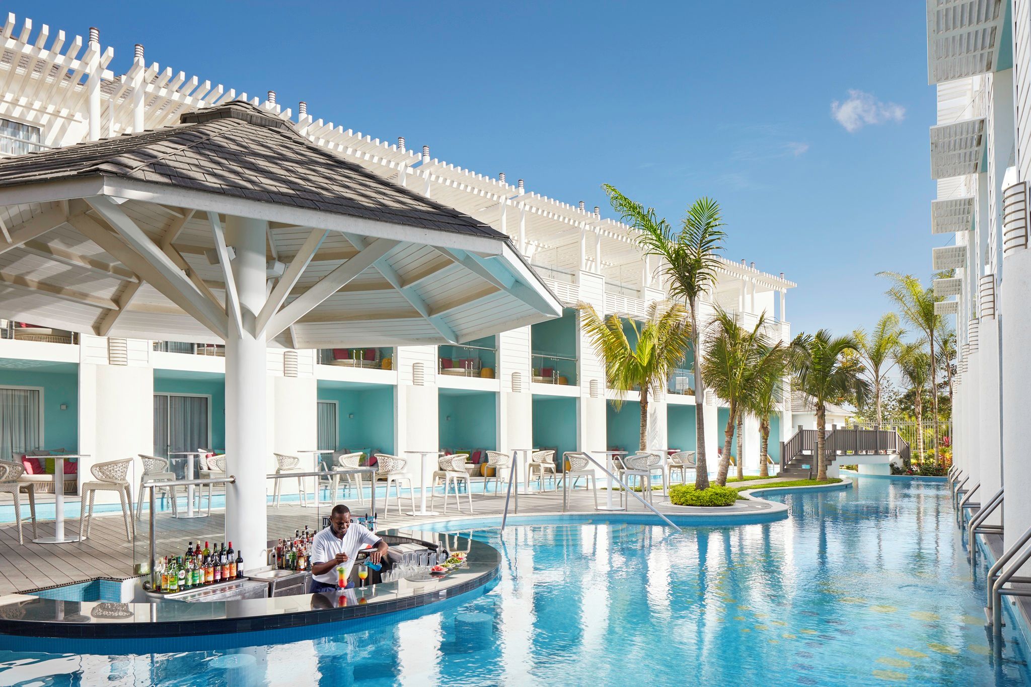 Uma área de bar à beira da piscina, com um bartender, espreguiçadeiras e piscina.