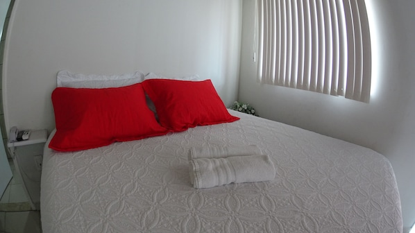 Cama bem arrumada com colcha branca e duas almofadas vermelhas, mesa de cabeceira branca com um telefone e uma janela com persianas.