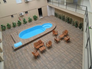 Área de piscina no terraço com piscina azul, cadeiras e mesas de madeira.