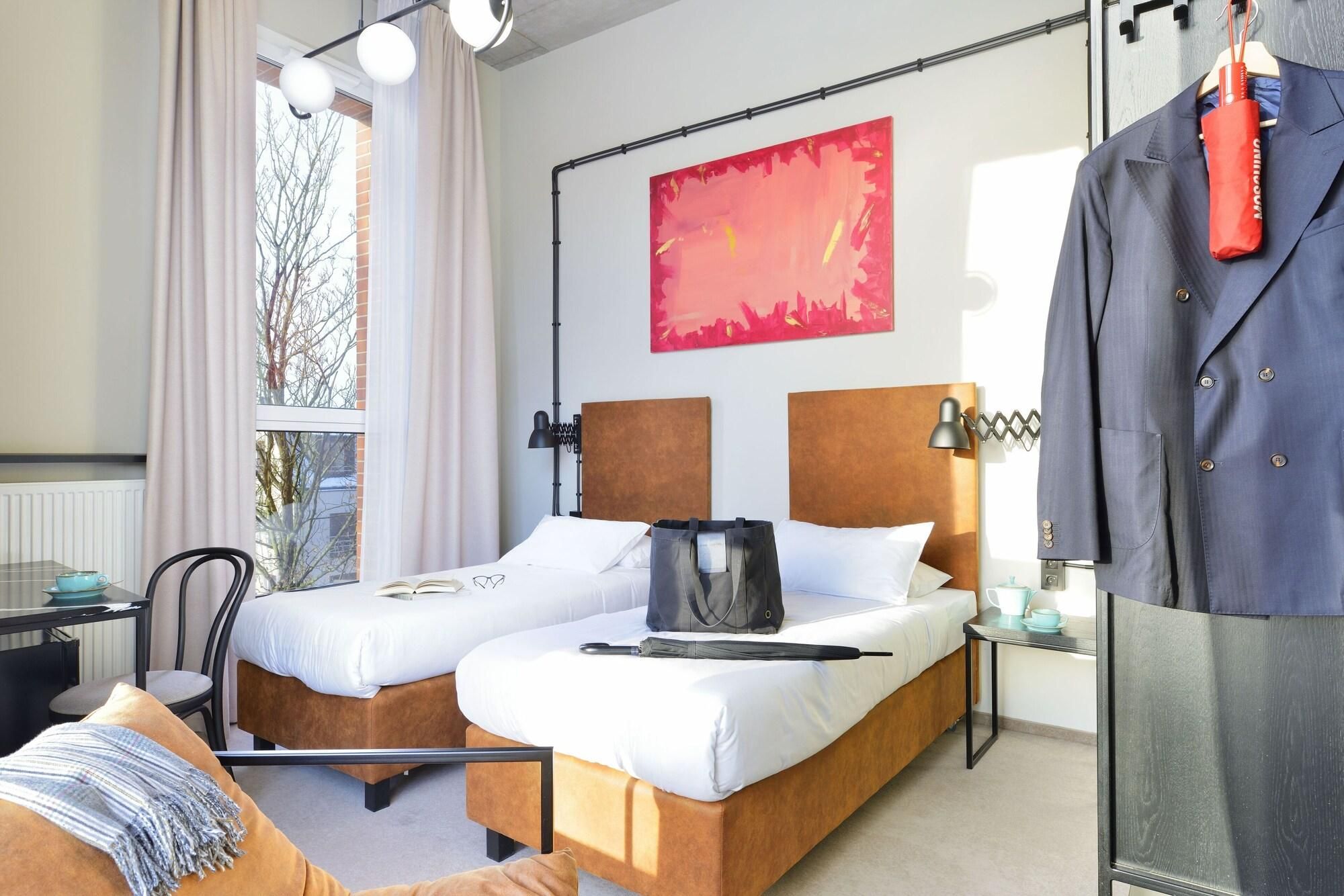 Quarto de hotel com duas camas, uma escrivaninha, uma cadeira, uma janela com cortinas, um quadro abstrato vermelho e um fato pendurado na parede.