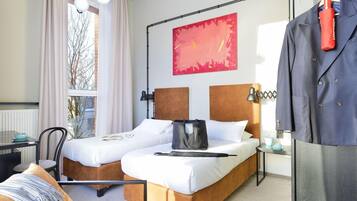 Quarto de hotel com duas camas, uma escrivaninha, uma cadeira, uma janela com cortinas, um quadro abstrato vermelho e um fato pendurado na parede.