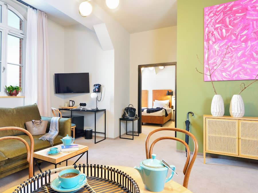 Uma sala moderna com sofá, mesa de centro, cadeiras, uma escrivaninha com computador e luminária, televisão e uma cama ao fundo.