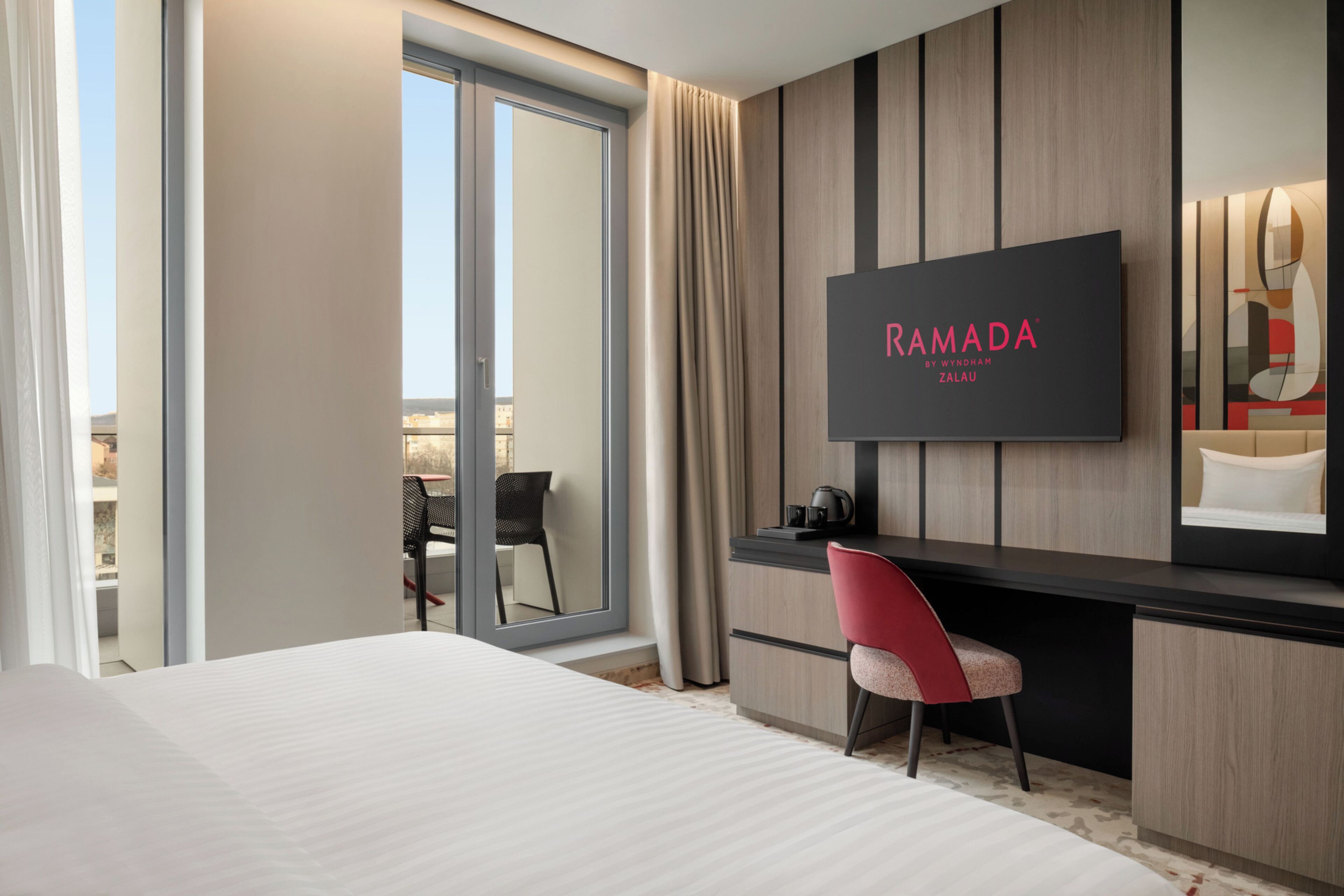 Quarto de hotel moderno com uma cama grande, uma mesa com cadeira, uma televisão fixada na parede e uma porta de vidro deslizante para uma varanda.