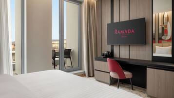 Quarto de hotel moderno com uma cama grande, uma mesa com cadeira, uma televisão fixada na parede e uma porta de vidro deslizante para uma varanda.