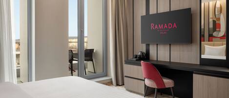 Quarto de hotel moderno com uma cama grande, uma mesa com cadeira, uma televisão fixada na parede e uma porta de vidro deslizante para uma varanda.