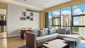 Uma sala de estar moderna com sofá seccional, uma pequena mesa redonda e um espaço de jantar com cadeiras. Janelas amplas oferecem vista para edifícios altos e áreas verdes.