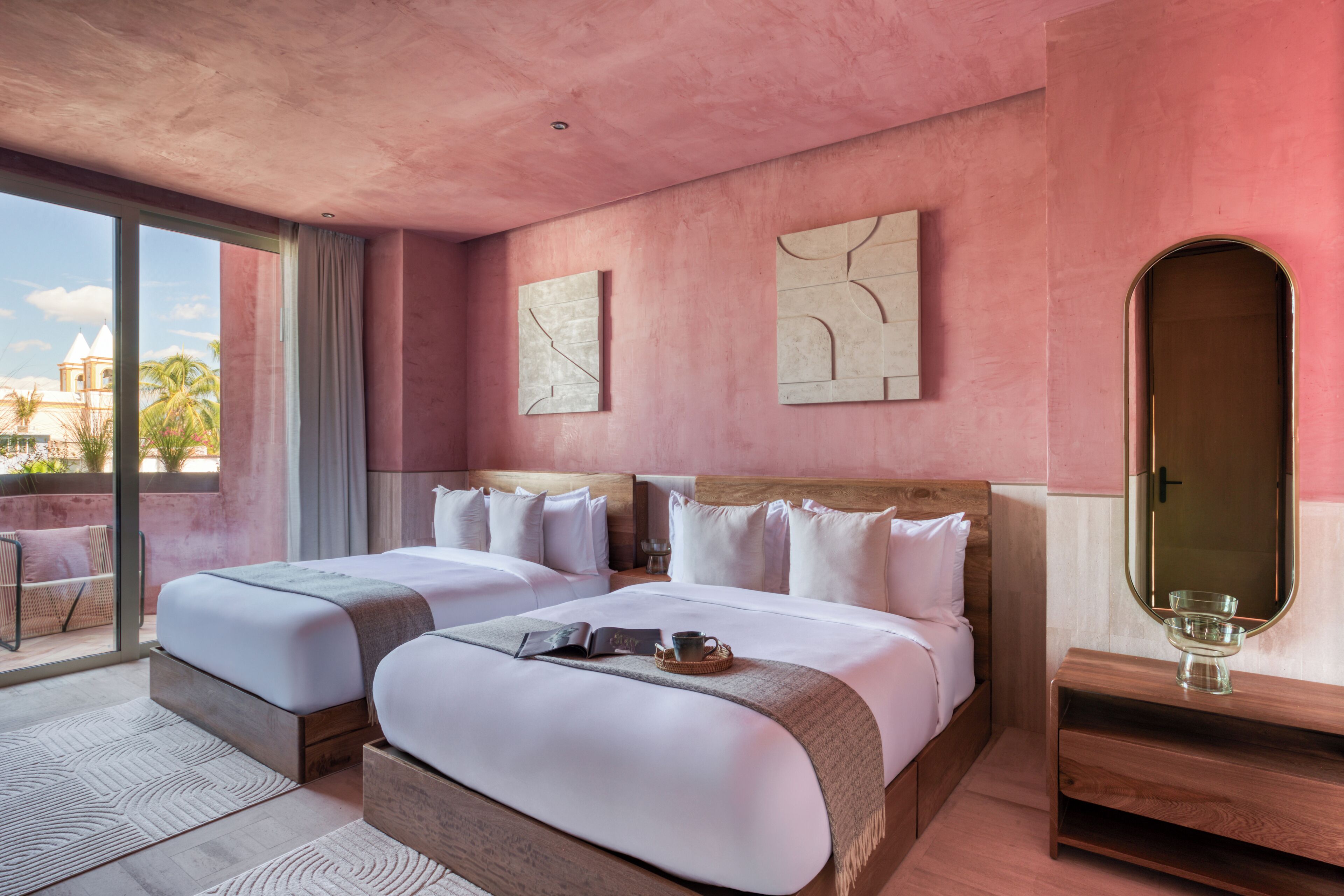 Quarto de hotel com duas camas, uma janela grande, uma varanda e uma mesa de cabeceira de madeira.