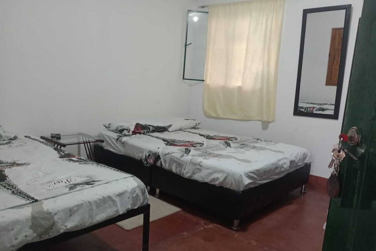 Quarto com duas camas de solteiro, uma mesinha, uma cadeira, um espelho e janela com cortinas.