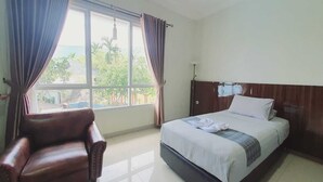 Quarto de hotel com uma cama, uma cadeira e uma janela grande com cortinas.