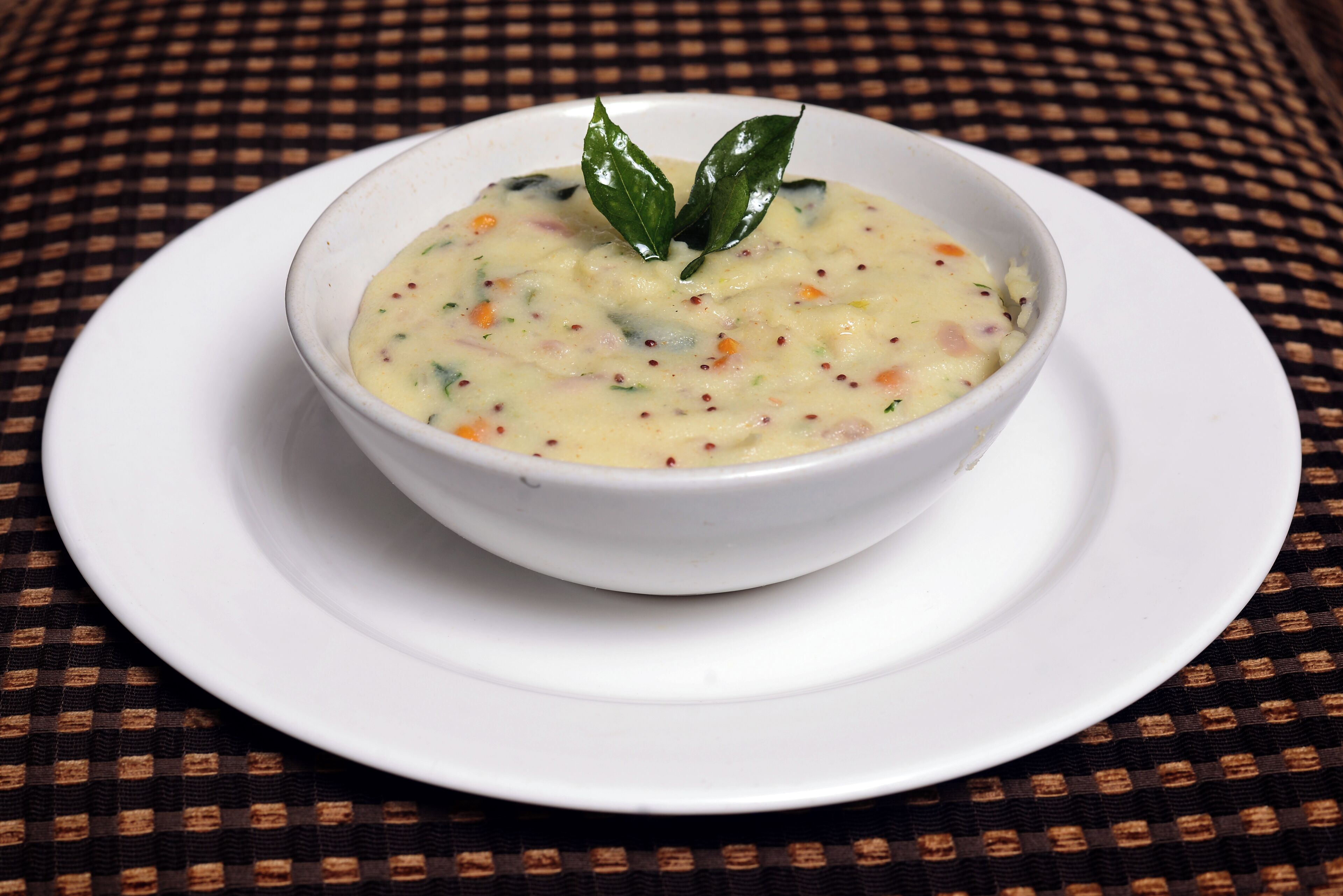 Uma tigela de sopa sobre um prato branco, com uma toalha de mesa quadriculada por baixo.