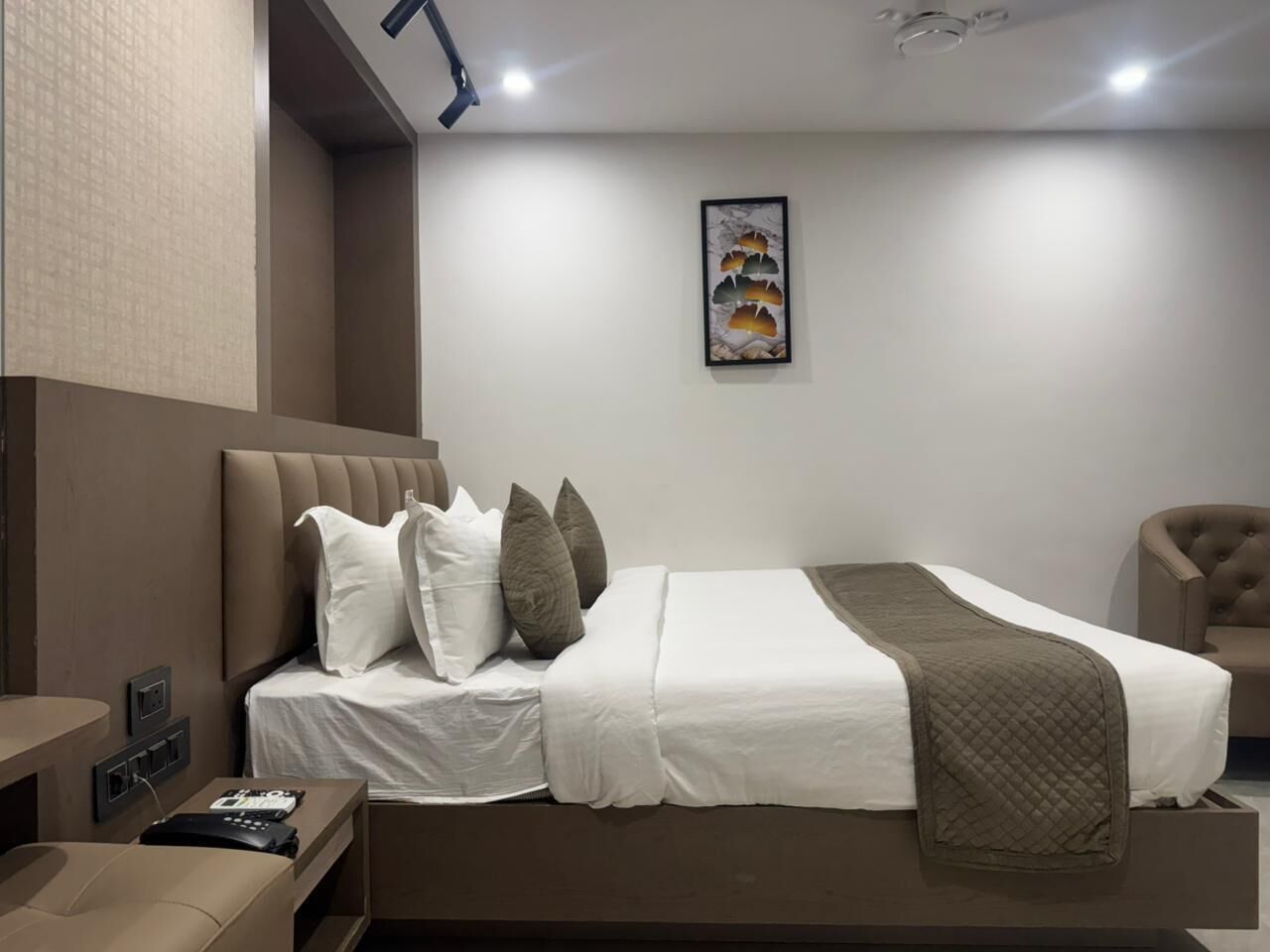 Quarto de hotel moderno com uma cama grande, mesa de cabeceira com telefone e controle remoto, uma poltrona e uma obra de arte pendurada na parede.