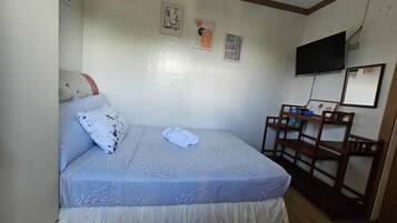 Quarto com cama, almofadas, televisão e escrivaninha com cadeira.