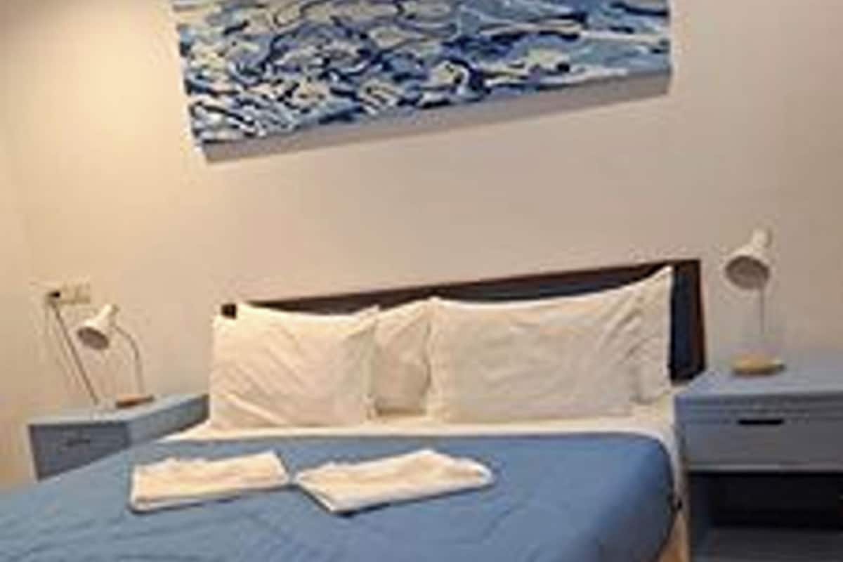 Uma cama bem arrumada com edredom azul e branco, uma luminária de cabeceira e uma mesinha com abajur.