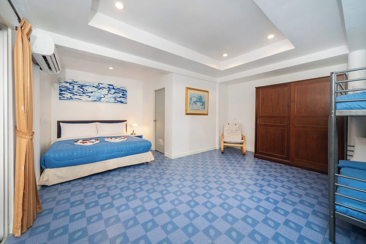 Um quarto com uma cama beliche, uma cama com colcha azul, um guarda-roupa de madeira, um quadro na parede e um chão estampado.