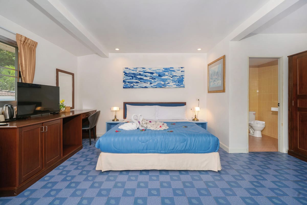Quarto de hotel com uma cama grande, uma mesa de trabalho com televisão e um banheiro visível através de uma porta aberta.