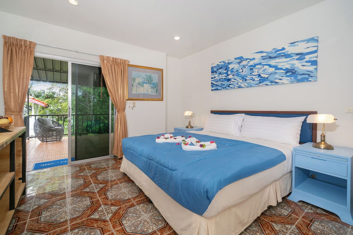 Quarto de hotel com cama, duas mesas de cabeceira, uma cadeira, varanda com cortinas e um quadro na parede.
