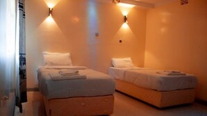 Quarto com duas camas, cada uma com roupa de cama branca e toalha dobrada. Há luminárias fixadas no teto.