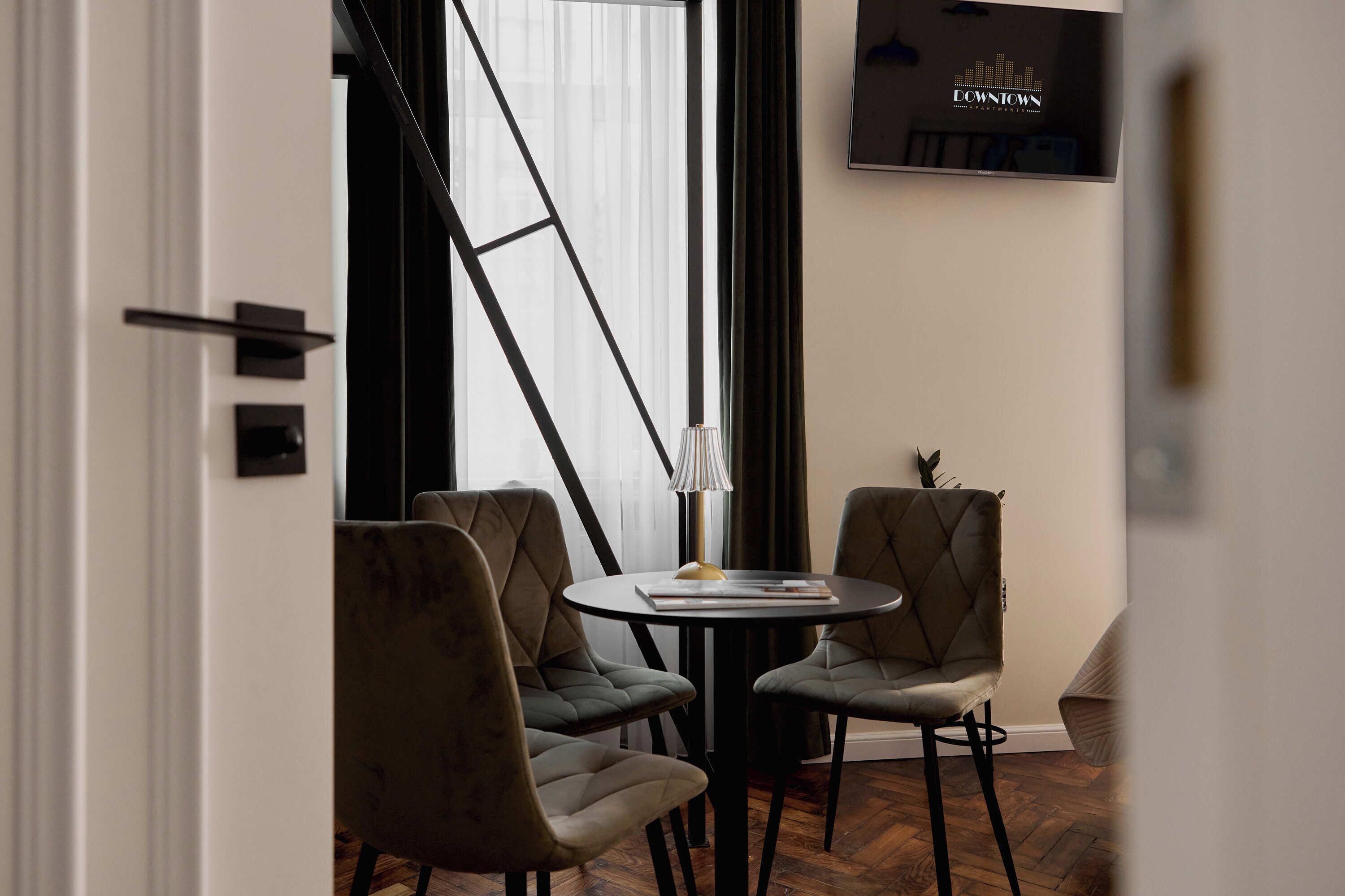 Interior moderno com área de refeições, apresentando duas cadeiras estofadas e uma pequena mesa redonda, uma escada de metal preta e uma televisão na parede exibindo 'DOWNTOWN APARTMENTS'.