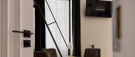 Interior moderno com área de refeições, apresentando duas cadeiras estofadas e uma pequena mesa redonda, uma escada de metal preta e uma televisão na parede exibindo 'DOWNTOWN APARTMENTS'.