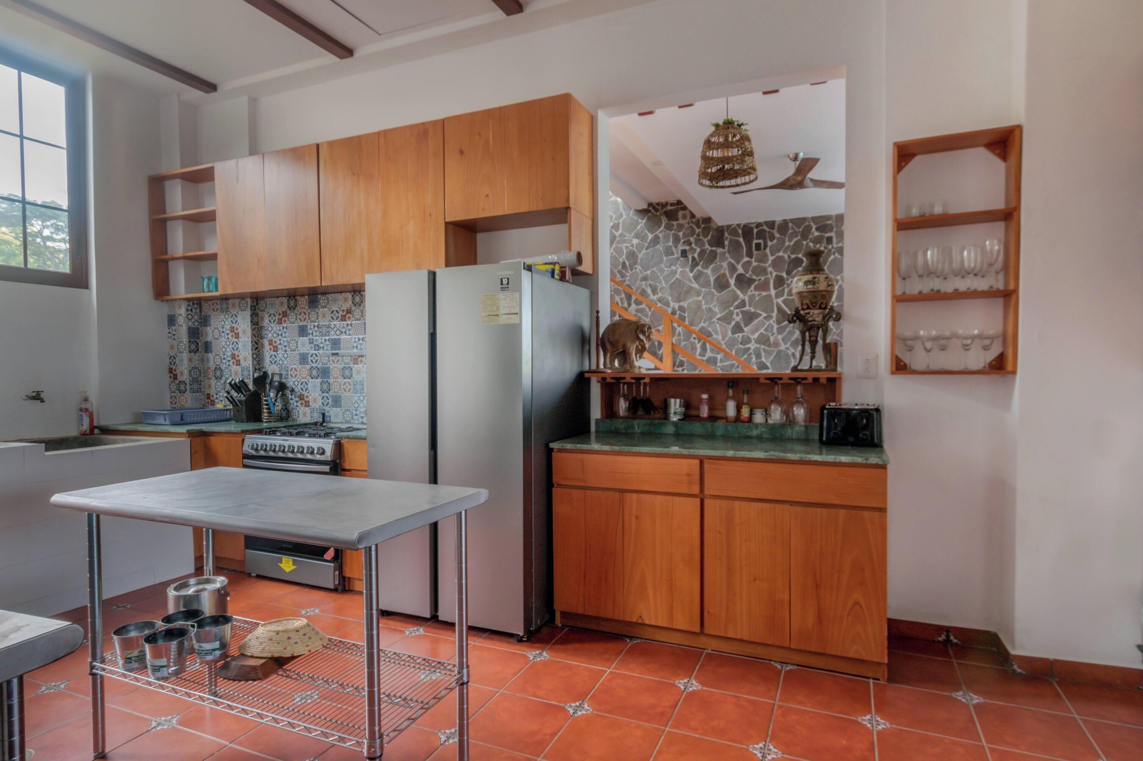 Cozinha com armários de madeira, uma geladeira de aço inoxidável, painel de azulejos e uma pequena mesa de jantar com copos de metal.