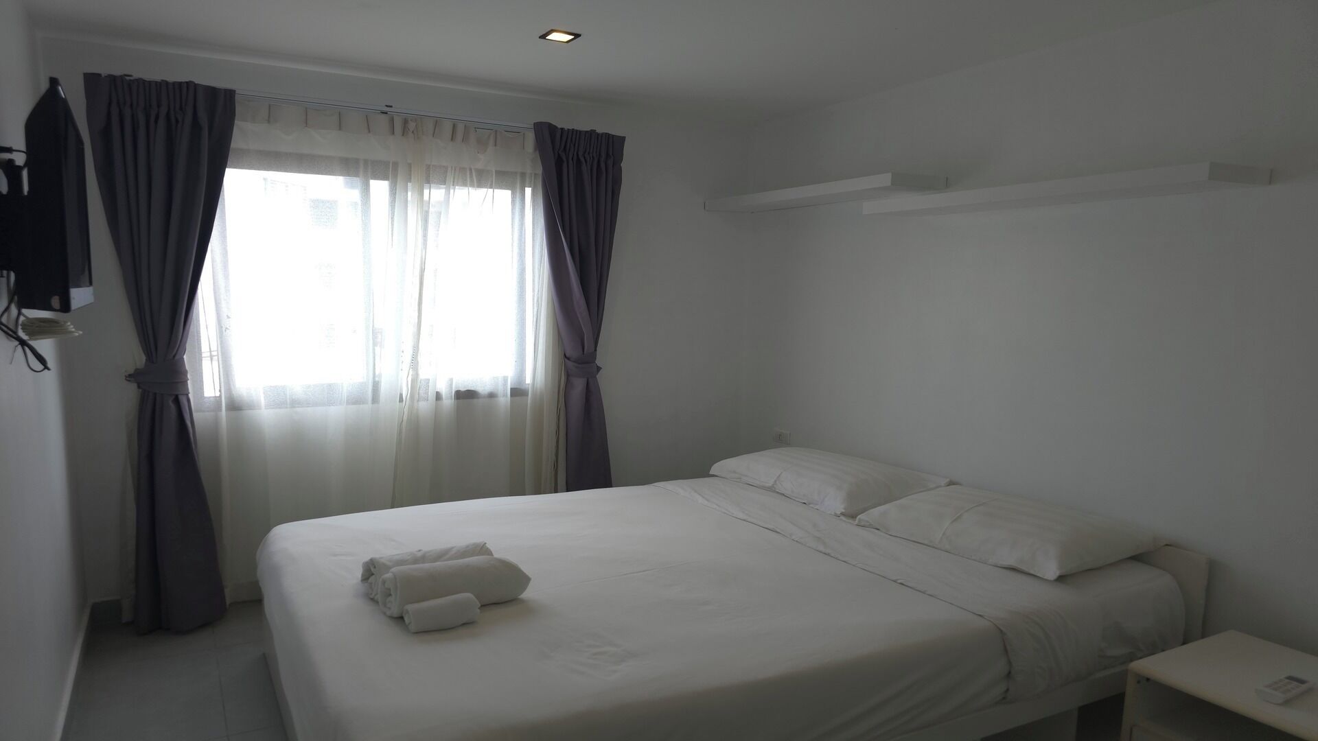 Quarto individual com uma grande janela, cortinas brancas, mesa de cabeceira e televisão de tela plana fixada na parede.