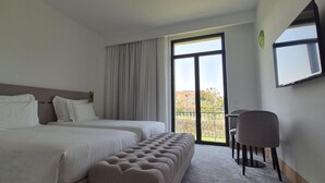 Quarto de hotel com duas camas, uma janela grande, uma cadeira e vista para o exterior.