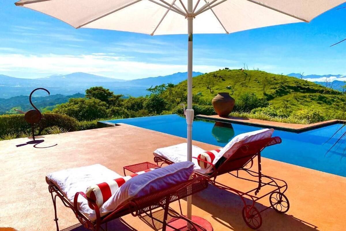 Área junto à piscina com duas espreguiçadeiras e um guarda-sol, com vista para uma paisagem pitoresca com colinas e céu claro.