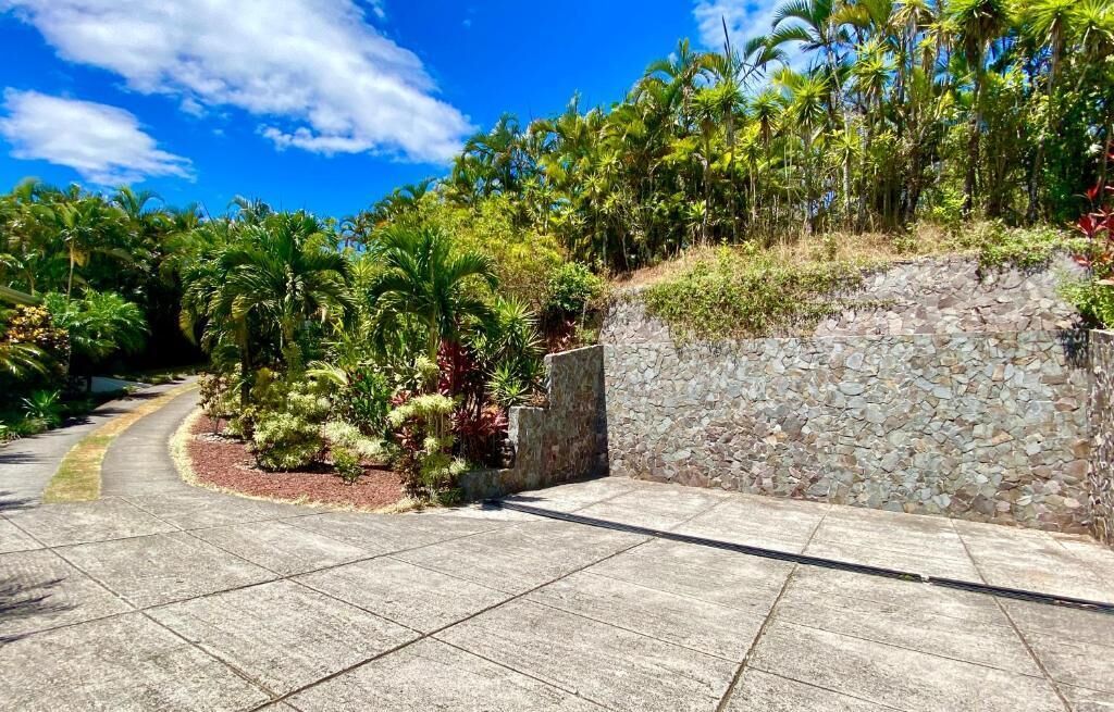 Um caminho pavimentado que leva a um muro de pedra, com vegetação tropical de um lado e um céu azul claro acima.