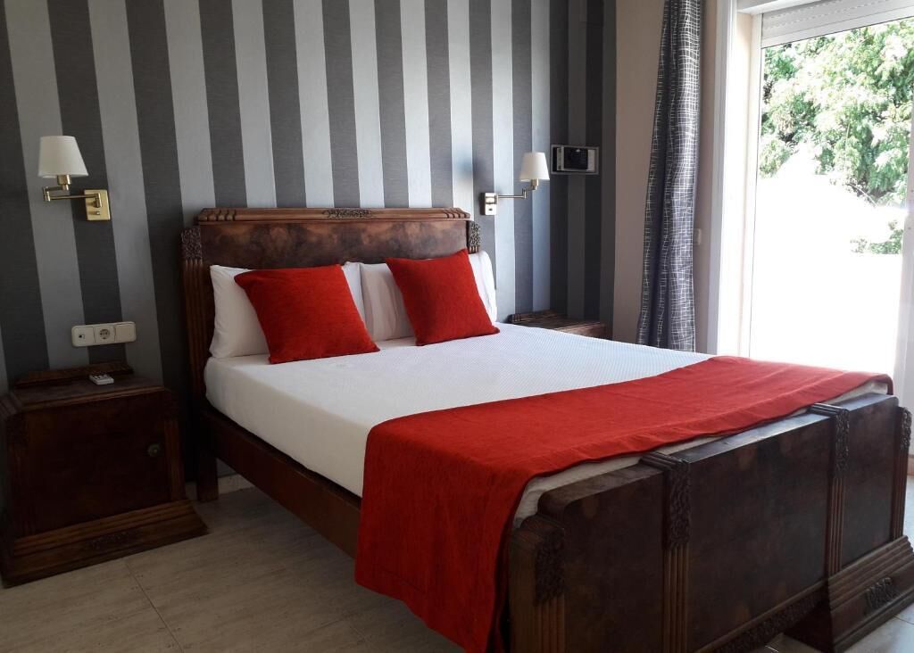 Uma cama bem arrumada com cabeceira de madeira, dois travesseiros vermelhos e um cobertor vermelho. Há duas mesas de cabeceira com abajures e uma TV fixada na parede.