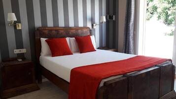 Uma cama bem arrumada com cabeceira de madeira, dois travesseiros vermelhos e um cobertor vermelho. Há duas mesas de cabeceira com abajures e uma TV fixada na parede.
