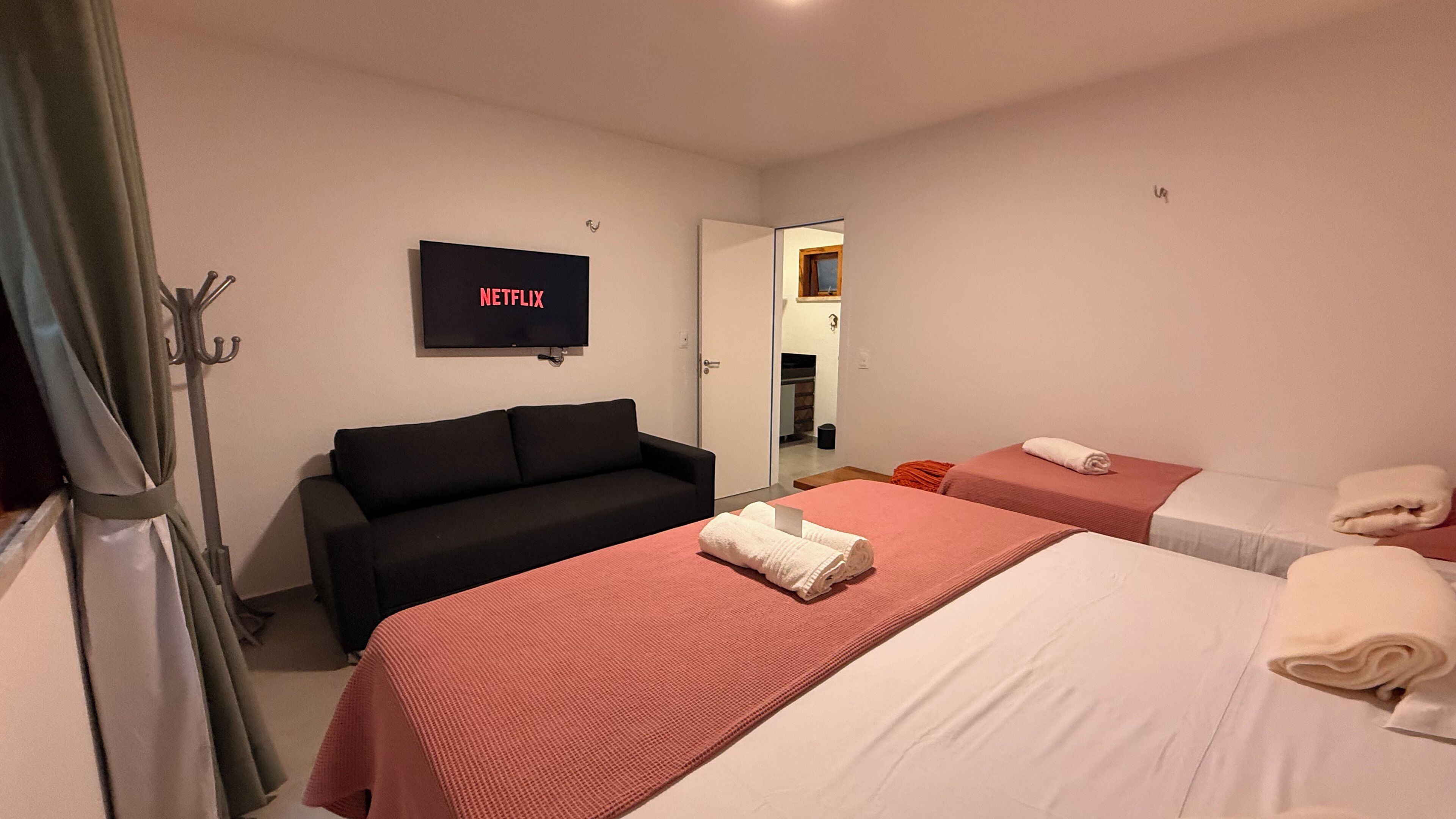 Quarto de hotel com duas camas, um sofá preto, uma TV exibindo 'NETFLIX' e um banheiro visível por uma porta aberta.