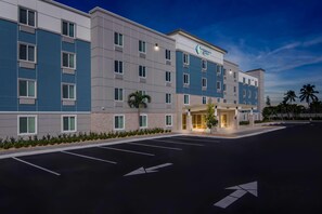 Hotel com vários andares, fachada moderna, entrada bem iluminada e estacionamento com setas indicativas.