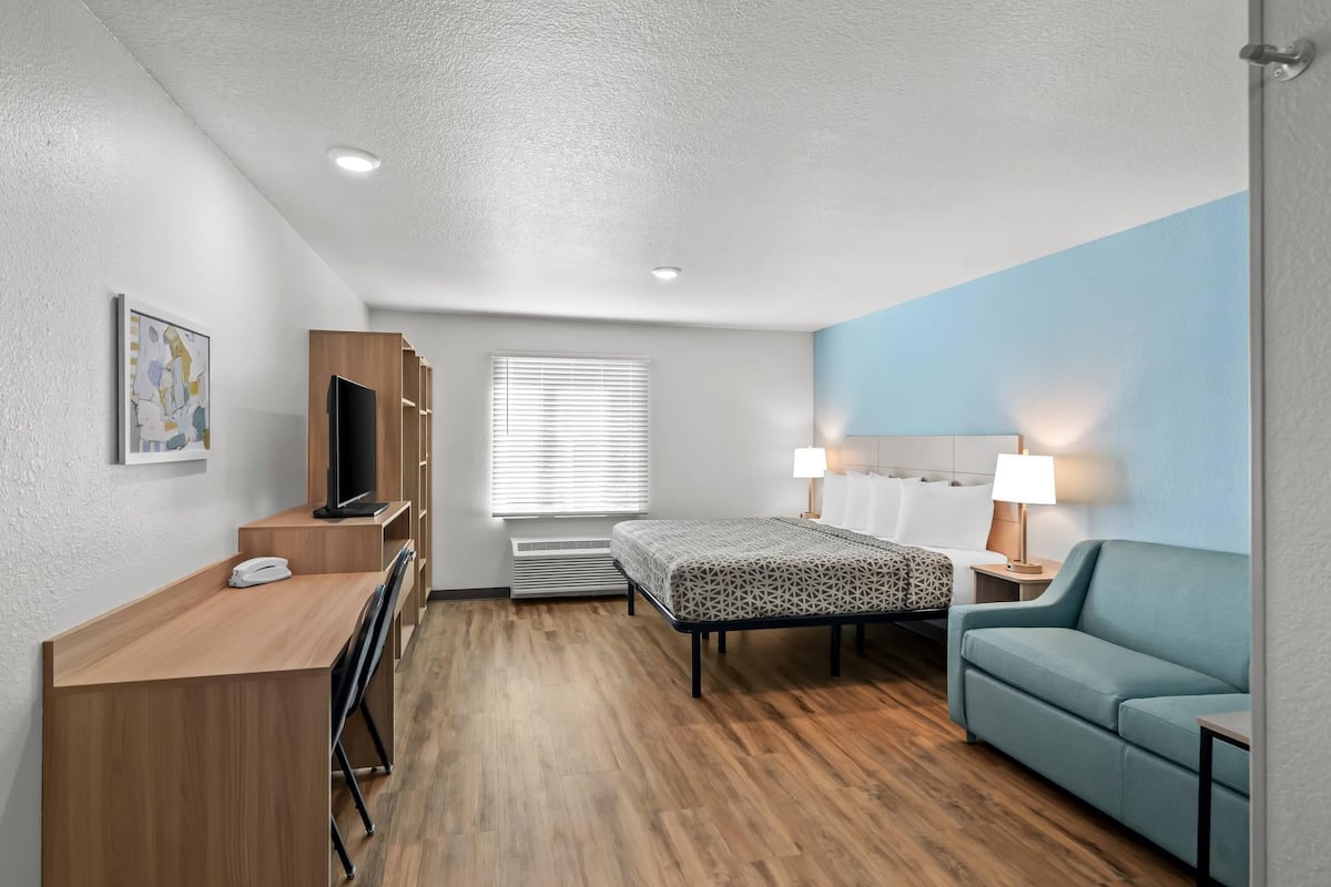 Quarto de hotel compacto com uma secretária de madeira, uma cama com roupa de cama estampada, um sofá azul e uma televisão.