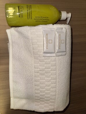 Uma toalha branca com padrão texturizado, um frasco verde de shampoo e dois pacotes brancos com a etiqueta "RESIDENZIALE".