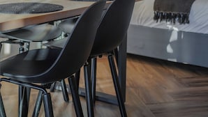 Quarto de hotel moderno com mesa de jantar de madeira, cadeiras pretas, cama com almofadas estampadas e um ventilador de teto.