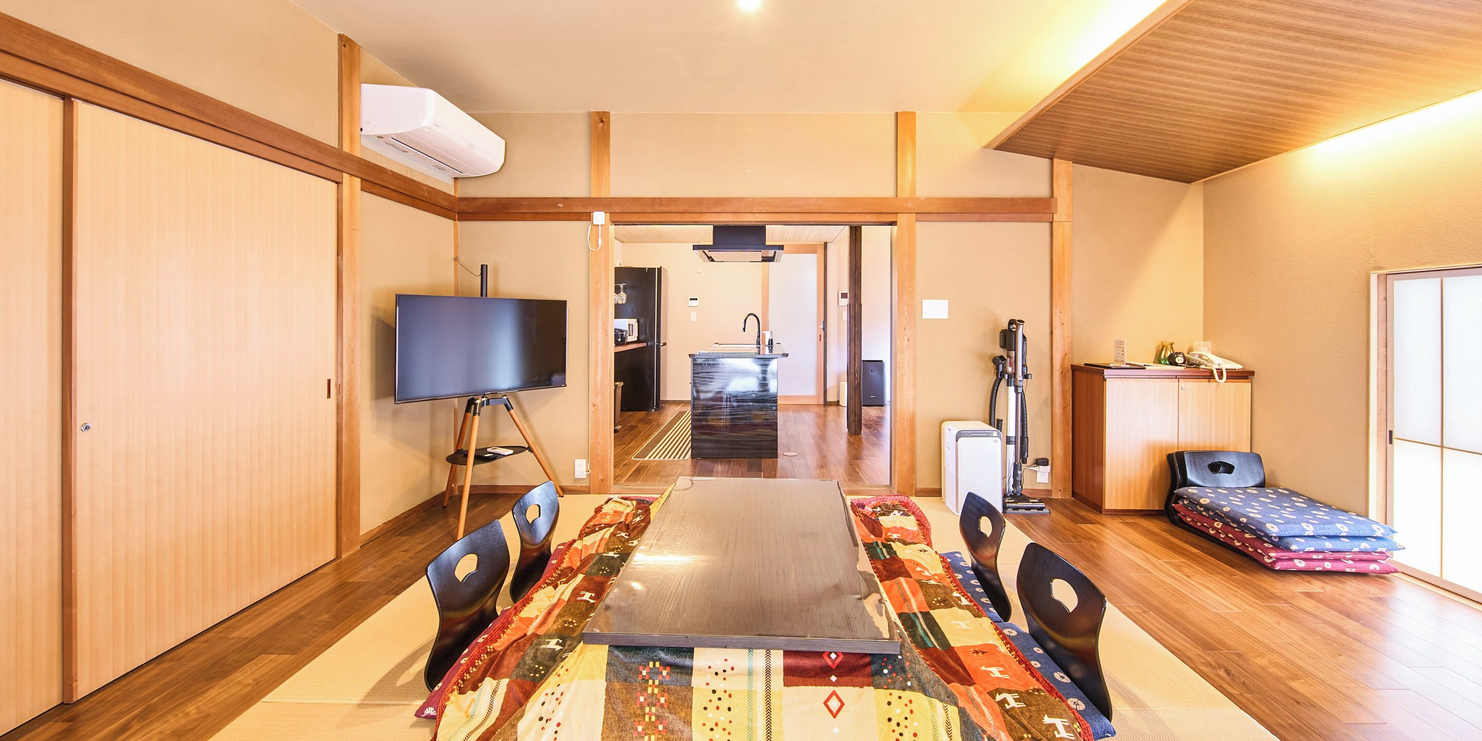 Um quarto tradicional japonês, com piso de tatame, uma mesa baixa com almofadas, uma televisão em um suporte de tripé e uma porta deslizante que dá para uma cozinha.