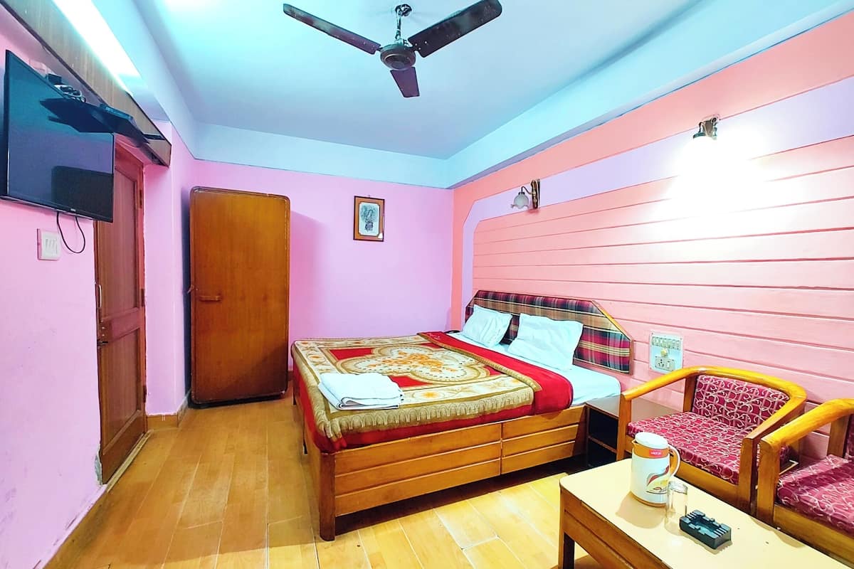 Quarto com cama, guarda-roupa de madeira, ventoinha no teto, televisão e mesa de cabeceira com uma chávena e um comando.
